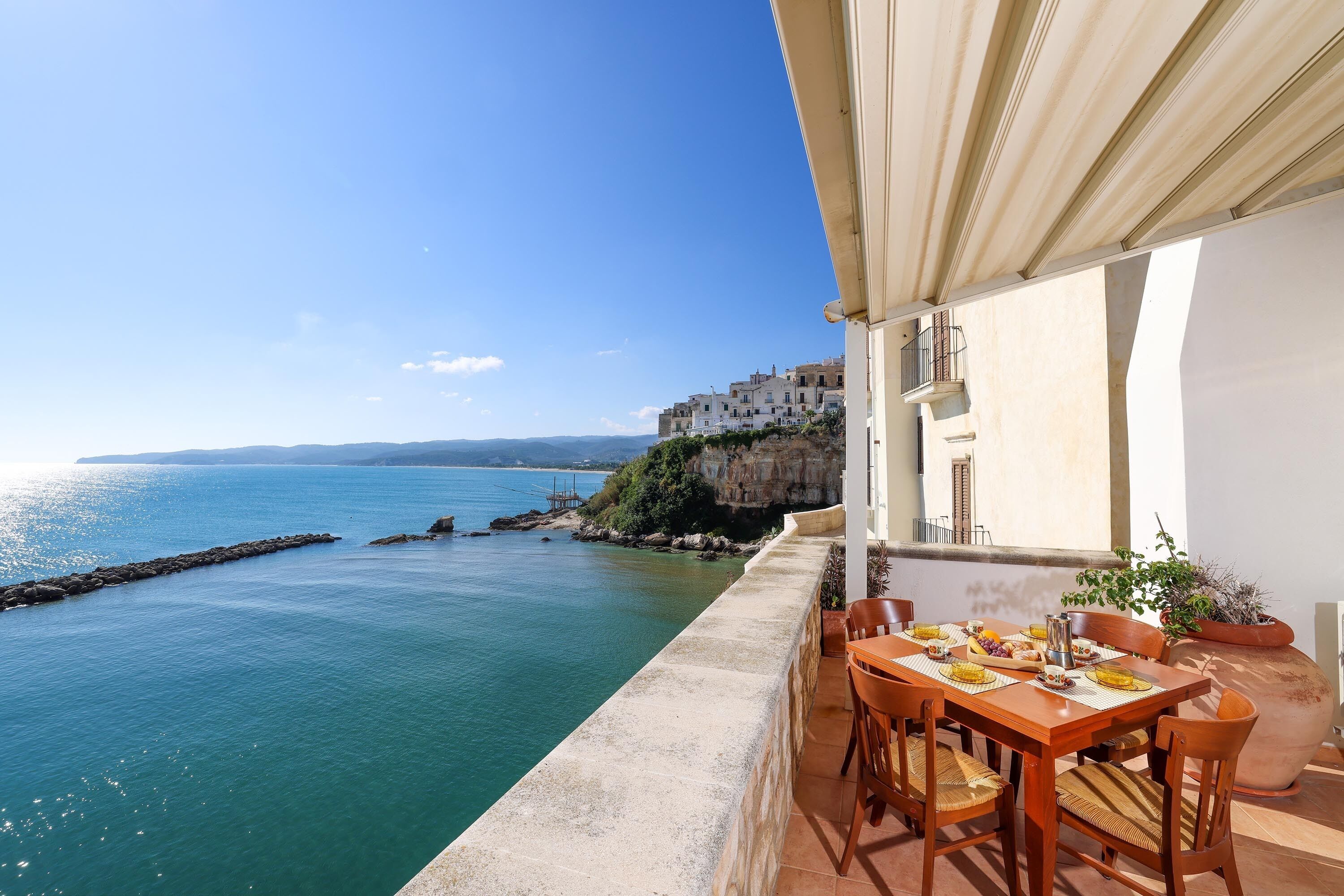 Terraço com mesa posta para refeições, com vista para a costa e edifícios em um penhasco.