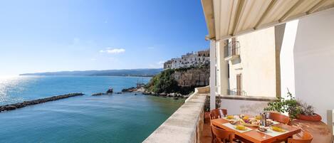 Terraço com mesa posta para refeições, com vista para a costa e edifícios em um penhasco.