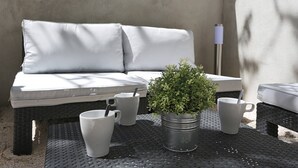 Uma área de pátio com um banco branco estofado, um vaso de metal com uma pequena planta e duas canecas brancas com colheres.