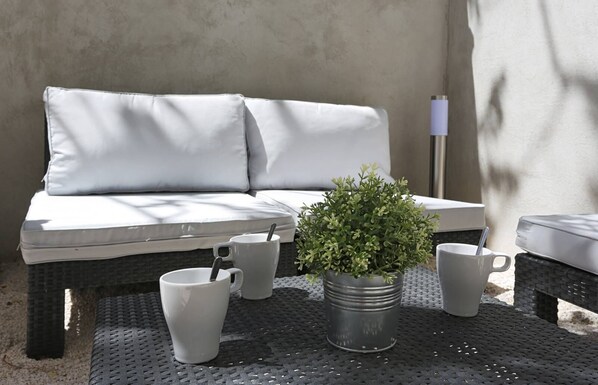 Uma área de pátio com um banco branco estofado, um vaso de metal com uma pequena planta e duas canecas brancas com colheres.