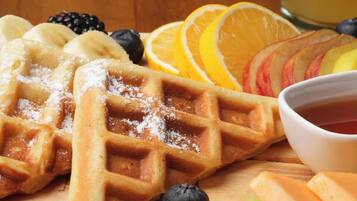 Um waffle com açúcar em pó, rodeado por frutas fatiadas e uma chávena de chá.