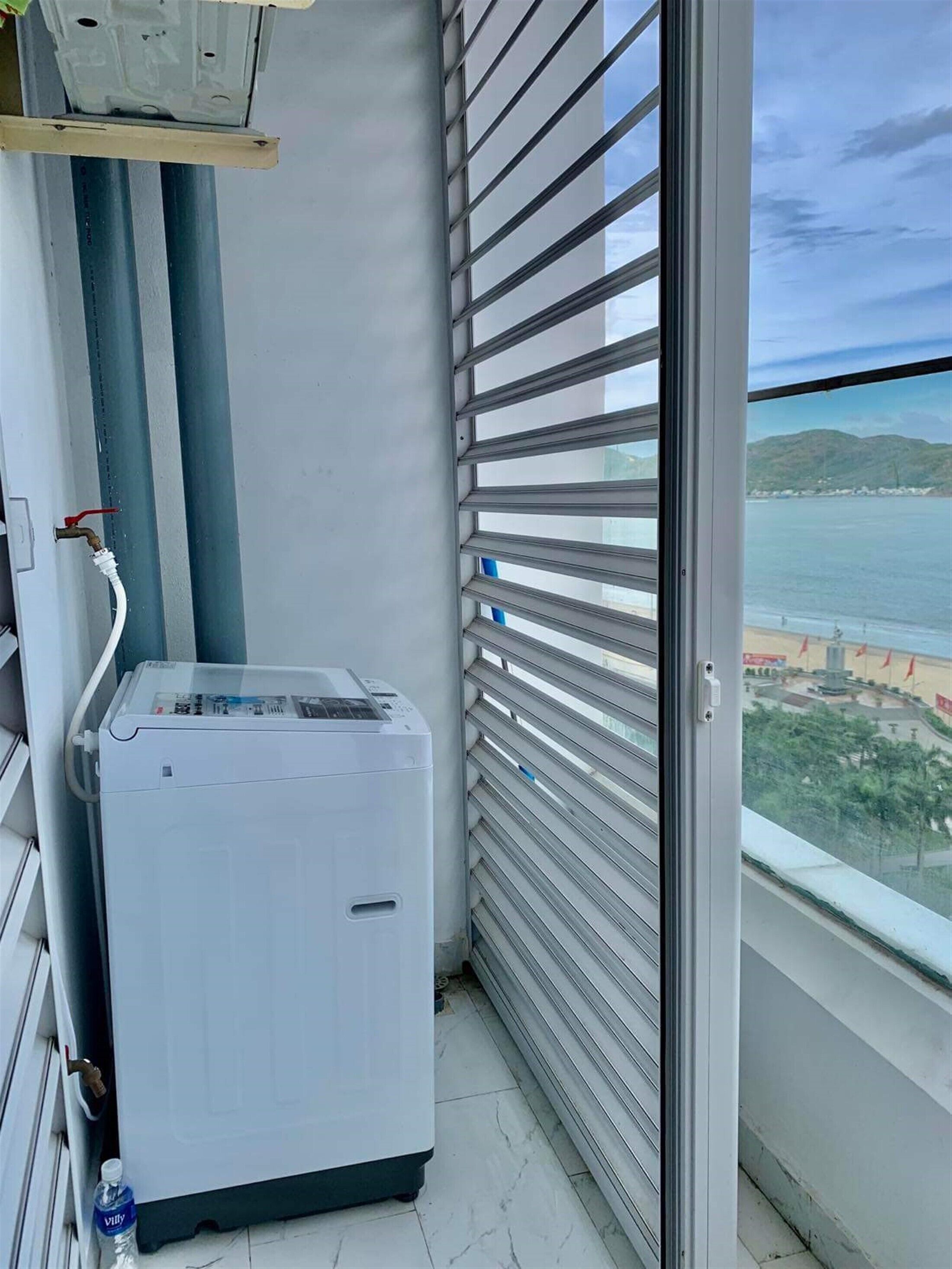 Uma pequena varanda com dispensador de água, uma janela com persianas horizontais e vista para a área costeira.