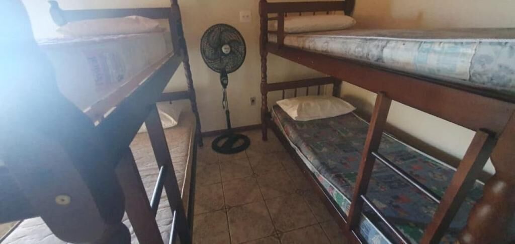 Quarto pequeno e compacto com beliches, uma mesa de madeira e um ventilador.
