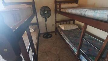 Quarto pequeno e compacto com beliches, uma mesa de madeira e um ventilador.