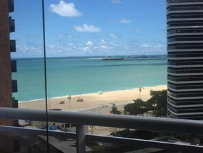 Vista de uma praia a partir de uma varanda, com céu claro, areal extenso e um navio grande ao longe.