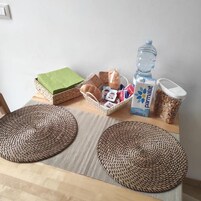 Uma mesa de madeira com jogos americanos trançados, um cesto de pão, uma garrafa de água, um pacote de leite Parmalat e um recipiente com aveia.
