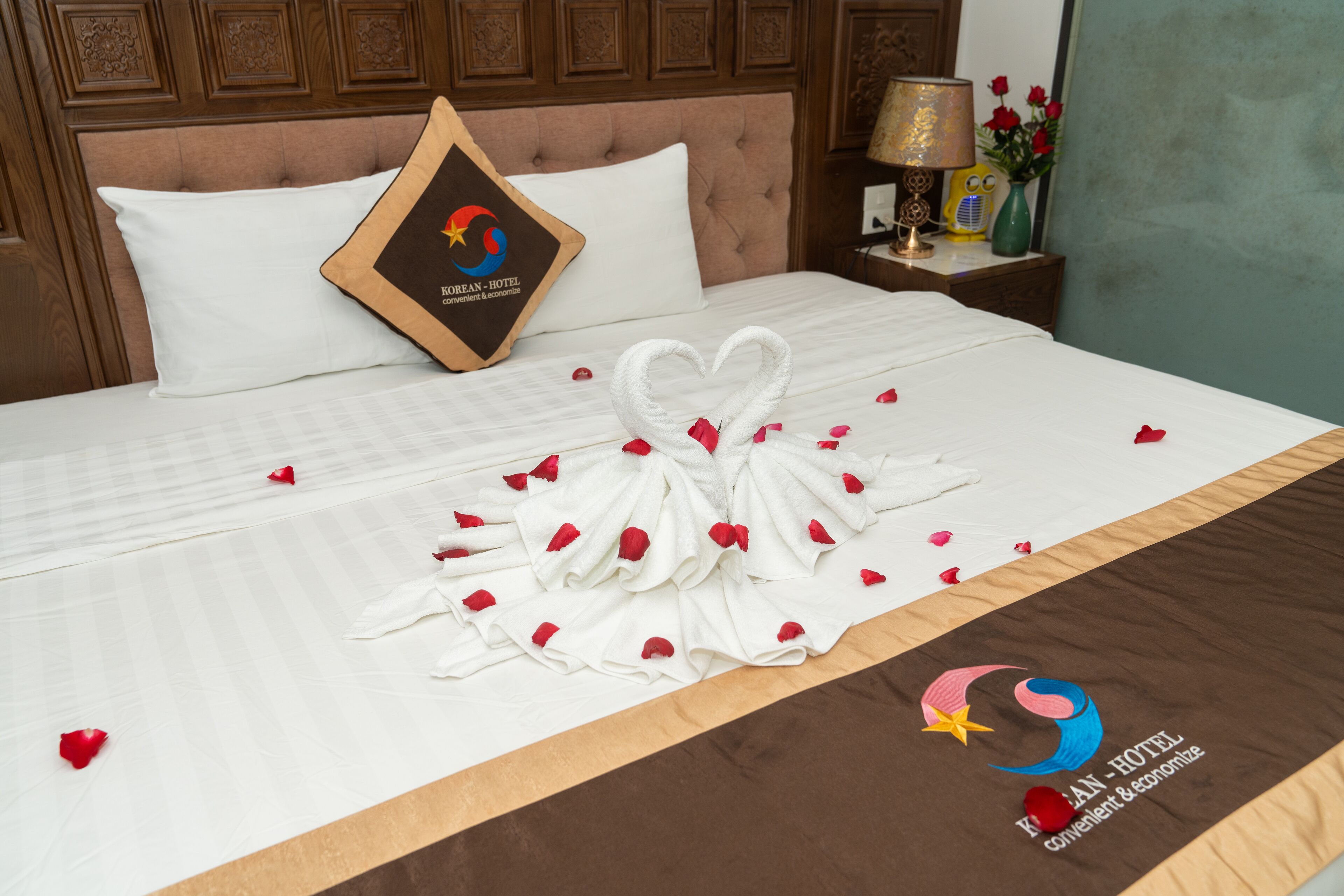 Uma cama bem arrumada com arranjo de toalhas em formato de coração e pétalas de rosa, um travesseiro com logotipo e o mesmo logotipo no cobreleito.