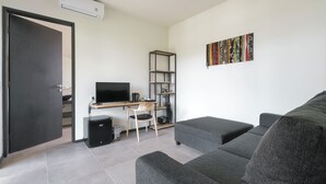 Uma sala de estar moderna com um sofá cinzento, uma TV de tela plana, uma cadeira de madeira e uma pequena escrivaninha com um computador.