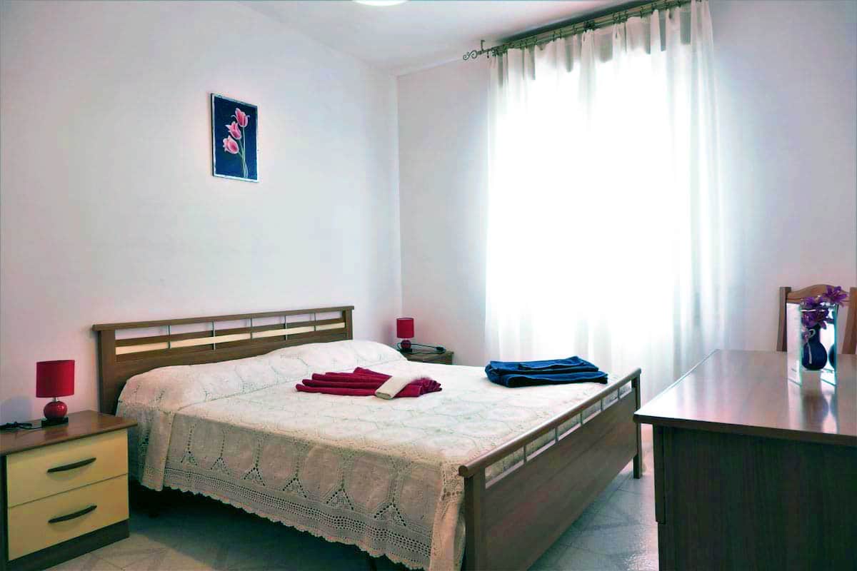 Quarto com cama, mesas de cabeceira, escrivaninha e janela com cortinas.