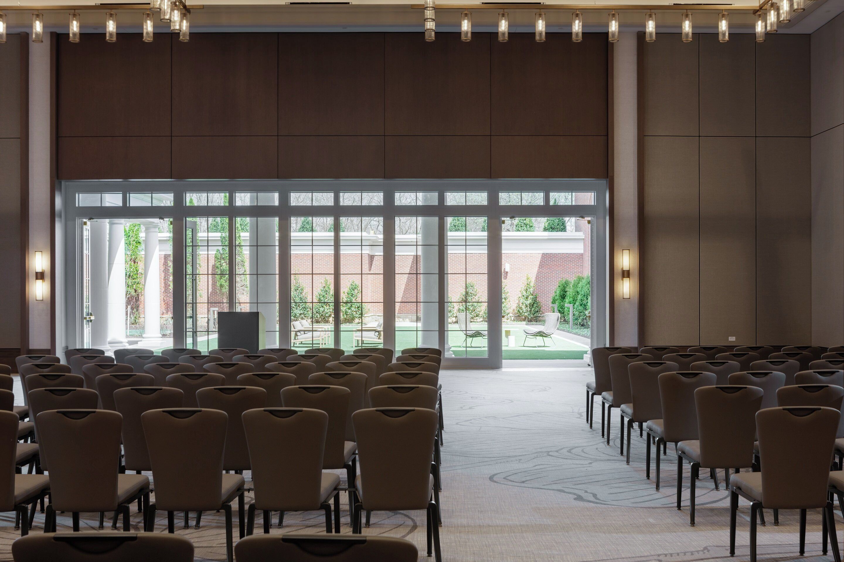 Uma sala de conferências com fileiras de cadeiras voltadas para uma janela grande, piso coberto por carpete e design moderno no teto.