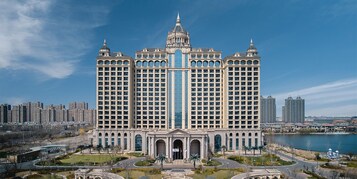 Um edifício grande, com vários andares e uma torre de vidro central, rodeado por construções menores e um corpo de água.