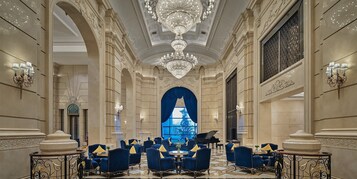 Um hall imponente com lustres ornamentados, um piano de cauda e poltronas azuis estofadas dispostas ao redor de uma mesa central.