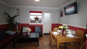 Um espaço de estar compacto com sofá, mesa de centro e mesa de jantar montados. Uma televisão está fixada na parede e é visível uma janela com vista para uma cerca.