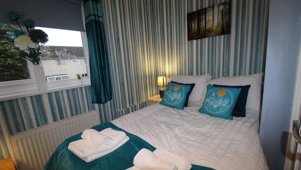Cama bem arrumada com edredom verde e branco, duas almofadas azul-esverdeadas e uma janela com vista para prédios.