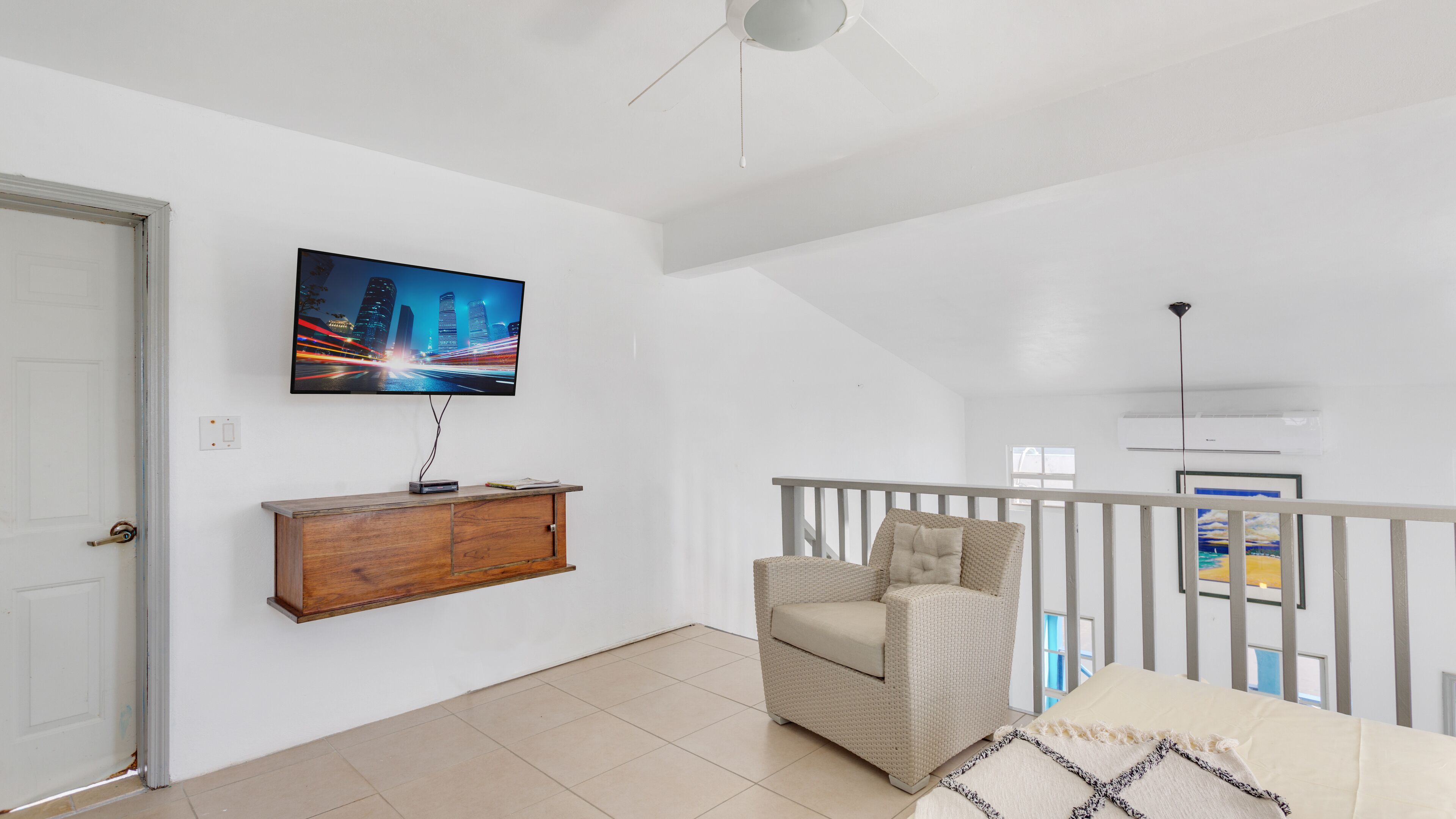 Uma sala de estar espaçosa com televisão de tela plana na parede, um armário de madeira abaixo, uma poltrona de vime e uma varanda com guarda-corpo.