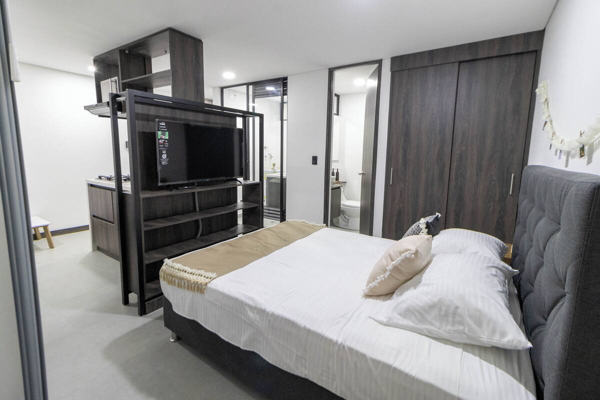 Quarto moderno com cama, televisão, guarda-roupa e banheiro visível por uma porta aberta.