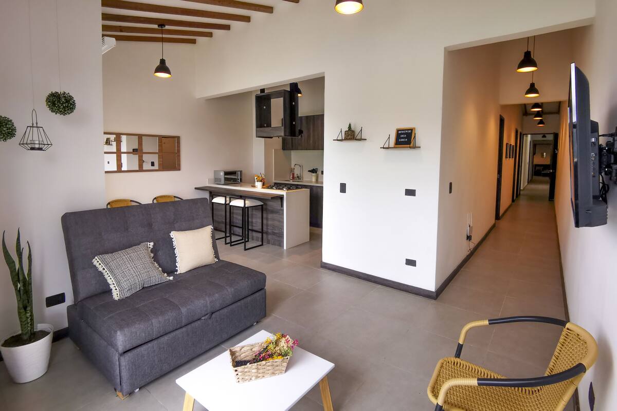 Uma sala de estar moderna com um sofá cinzento, uma mesa de centro branca e uma cadeira de vime. Dá para ver uma área de cozinha ao fundo, com uma mesa de jantar e banquetas.