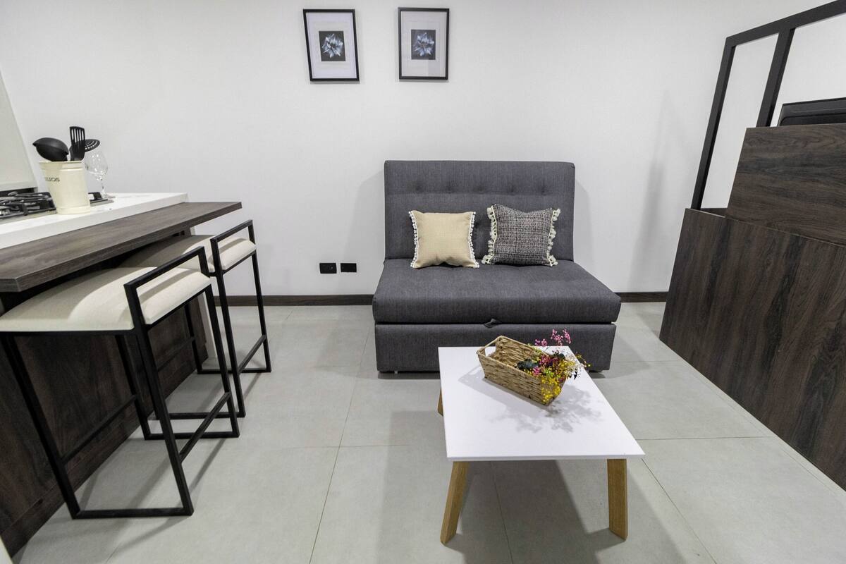 Uma sala de estar moderna com um sofá cinzento, uma mesa pequena branca e um cesto, além de uma cozinha com bancada e banquetas.