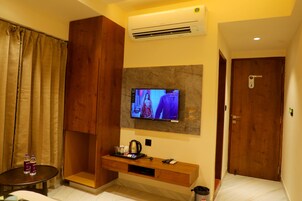 Quarto de hotel com televisão na parede, um armário de madeira, uma pequena mesa com chaleira e xícaras, e uma lixeira.