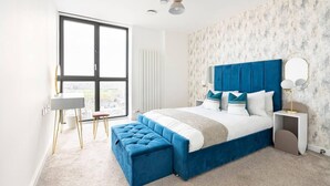Quarto moderno com uma grande janela, um banco estofado azul, uma mesa de cabeceira branca e uma cabeceira azul.