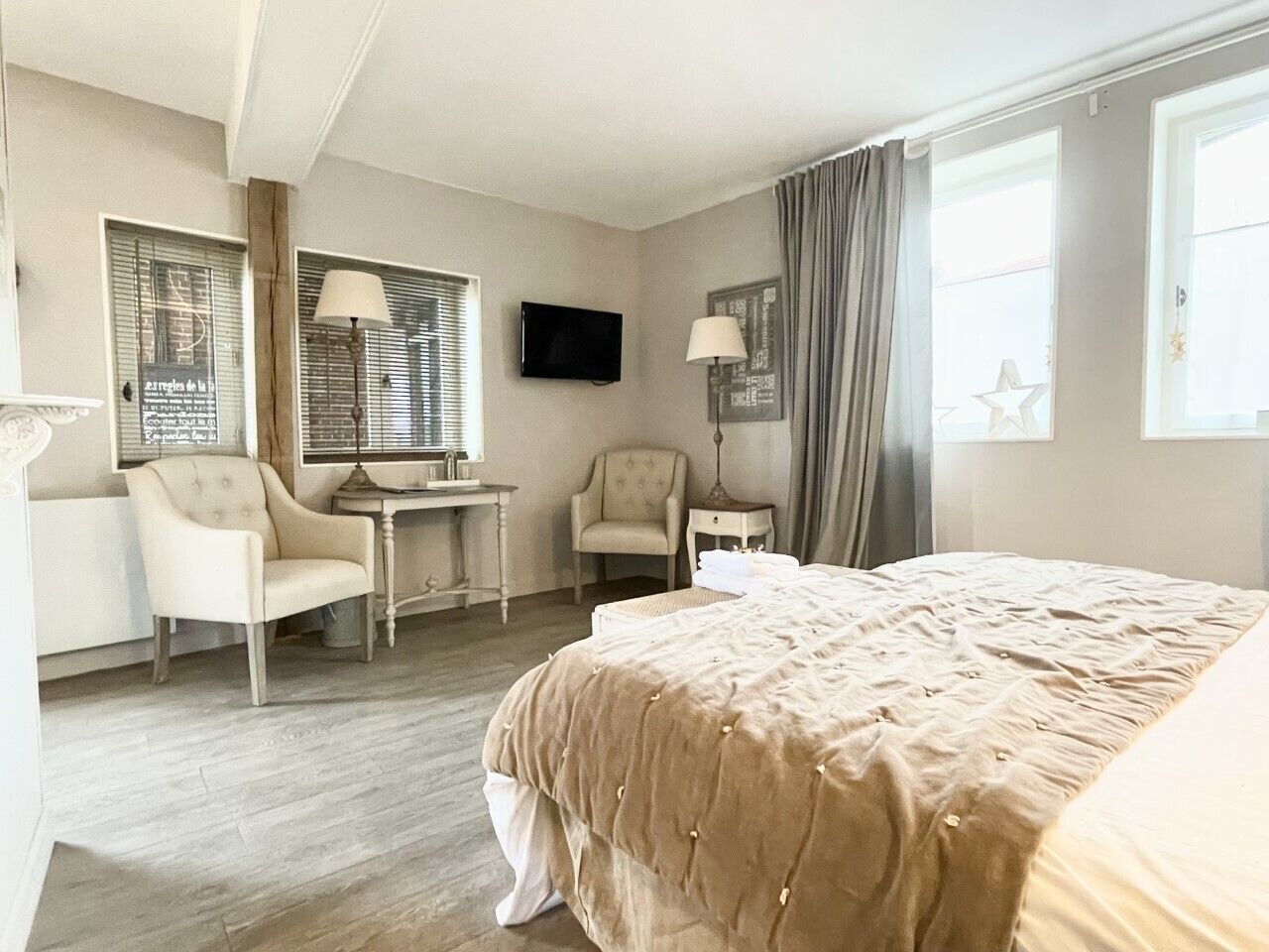 Quarto de hotel com uma cama grande, duas cadeiras, uma escrivaninha, uma televisão e uma janela com cortinas.