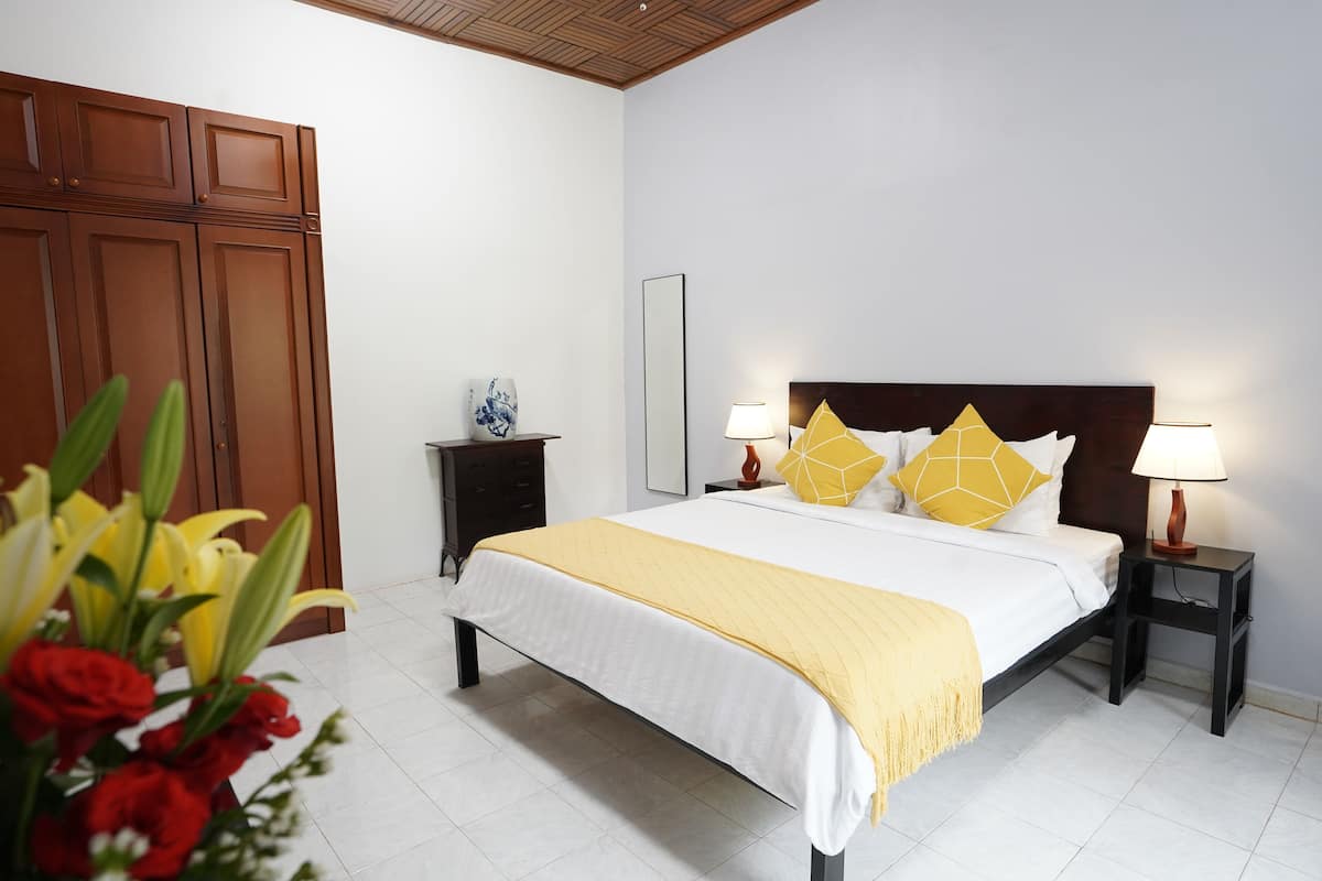 Uma cama bem arrumada com um edredom branco e amarelo, duas mesas de cabeceira com luminárias, um guarda-roupa de madeira e um espelho na parede.