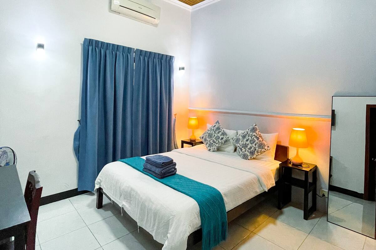 Quarto de hotel com cama, duas luminárias de cabeceira, ventilador de teto, janela com cortinas azuis e piso revestido com azulejos.