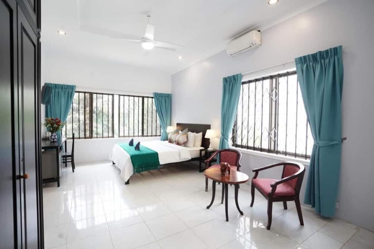 Quarto de hotel com cama, duas cadeiras, uma mesinha, escrivaninha com cadeira e janelas amplas com cortinas.