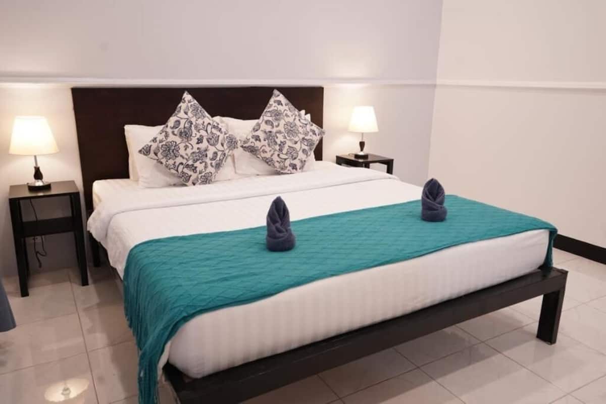 Cama bem arrumada com colcha verde-água e duas almofadas, com duas mesas de cabeceira com abajures, num quarto simples com paredes brancas e cabeceira escura.