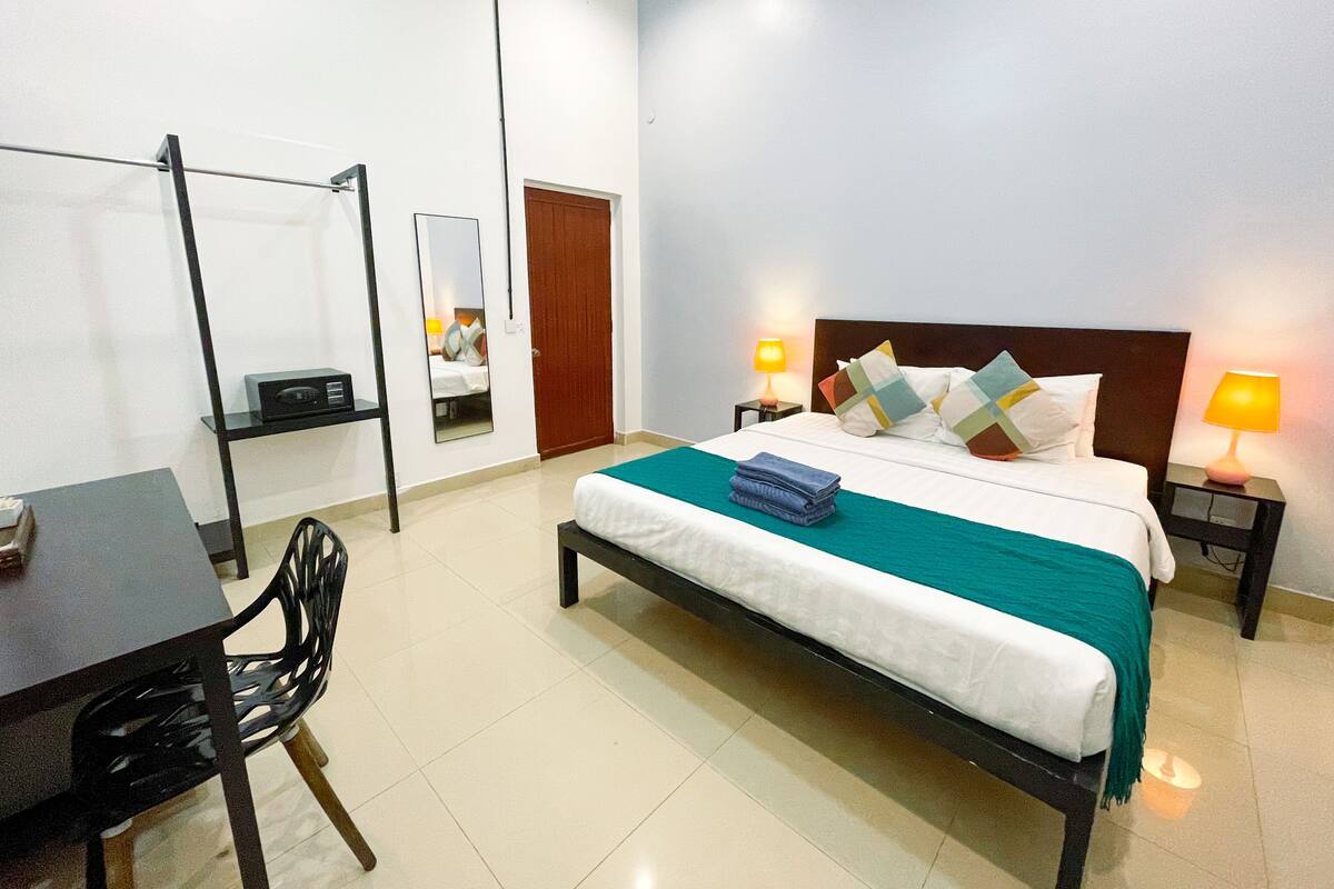 Quarto de hotel com cama, mesas de cabeceira, escrivaninha, cadeira e espelho.