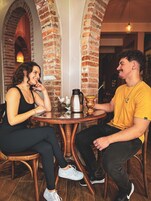 Duas pessoas sentadas numa mesa redonda, com uma cafeteira e uma chávena. O ambiente apresenta paredes de tijolo e mobiliário de madeira.