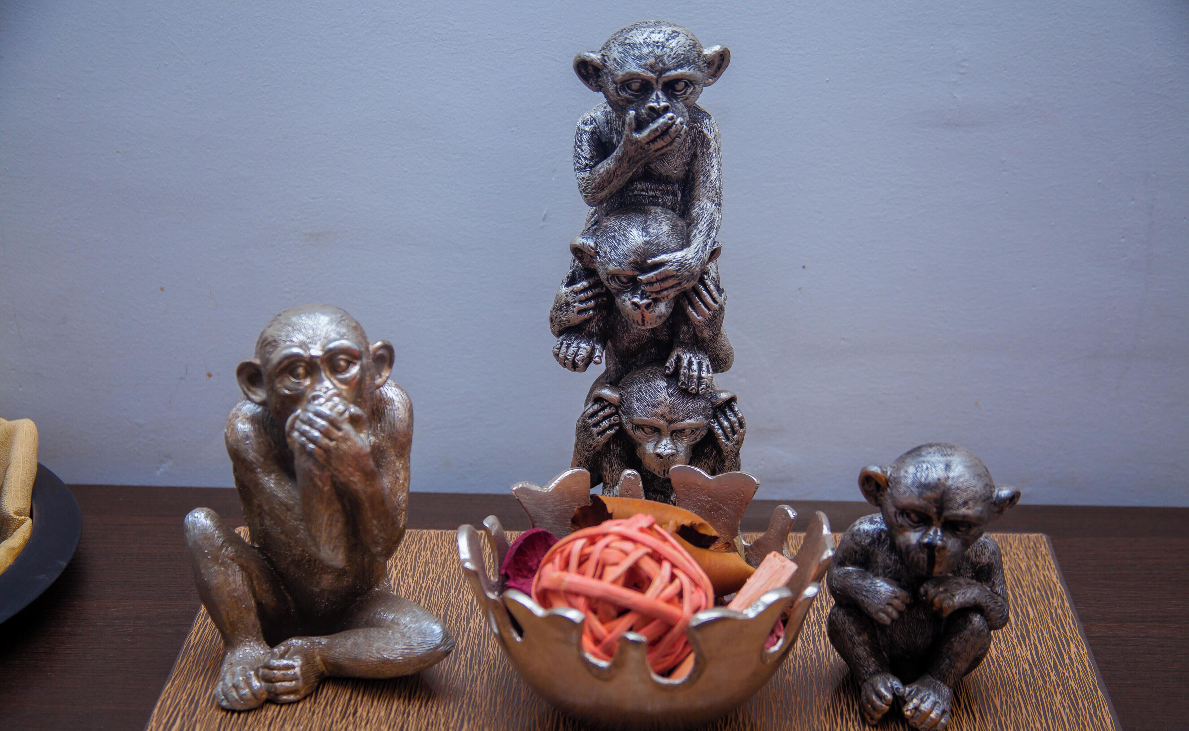 Três figuras de macaco sobre uma superfície de madeira, com uma tigela de objetos coloridos ao centro.