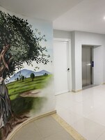 Corredor com um mural de uma árvore e paisagem, uma porta branca e um elevador moderno.