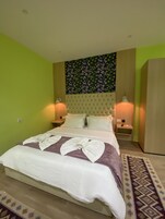 Cama bem arrumada com lençóis brancos e um cobertor roxo, duas luminárias de cabeceira, um painel de cama com padrão floral e uma parede verde.
