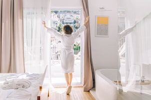 Quarto com cama, janela com cortinas, banheira e vista para edifícios lá fora.