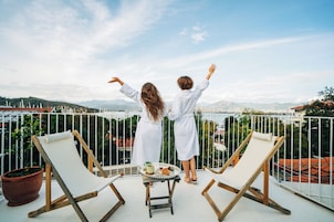 Uma varanda com duas poltronas de descanso, uma mesa com um croissant e uma xícara, e vista para montanhas e edifícios.