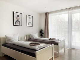 Quarto de hotel moderno com duas camas, uma mesa de cabeceira, um vaso de plantas e janelas amplas com cortinas leves.