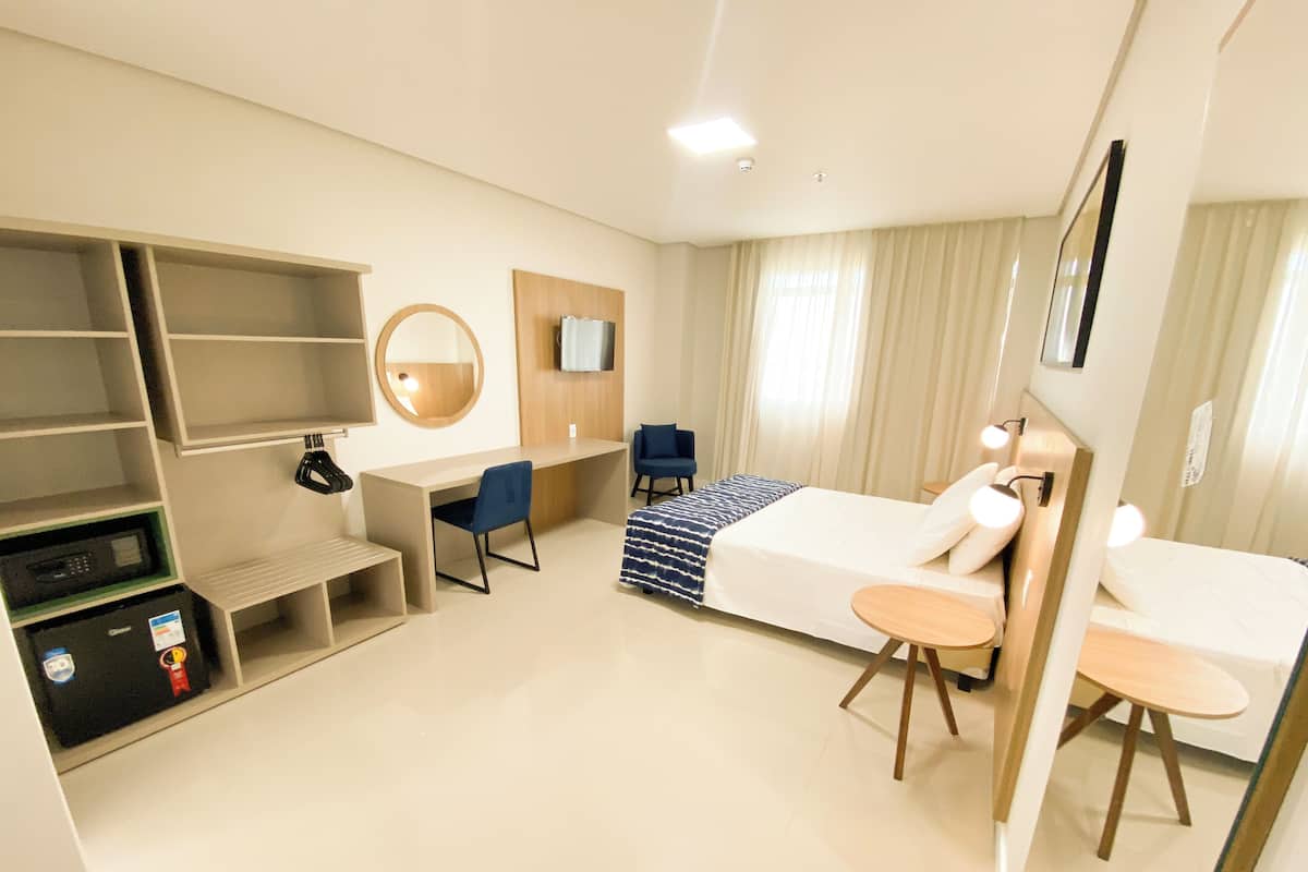 Quarto de hotel com cama, secretária, cadeira, televisão e guarda-roupa.