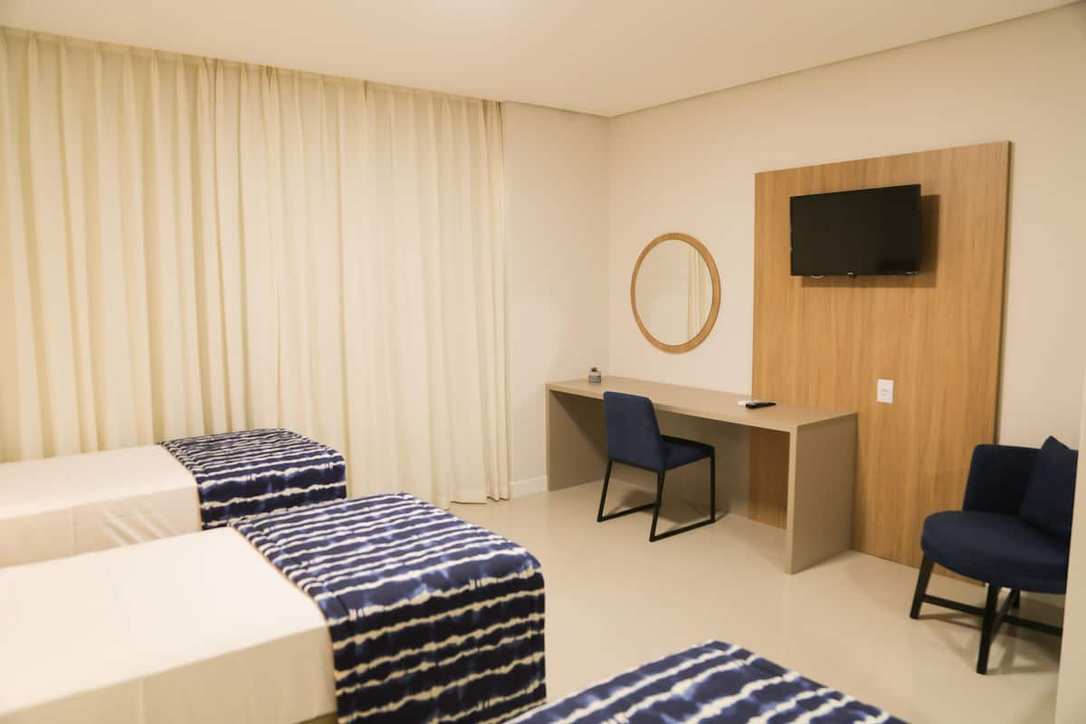 Quarto de hotel com cama, escrivaninha, cadeira e televisão fixada na parede.