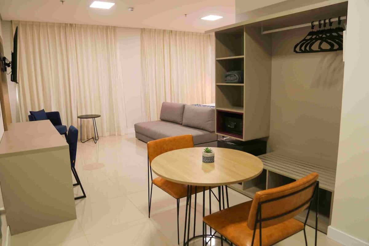 Uma sala de estar moderna com um sofá, uma mesa de jantar com cadeiras, uma mesinha lateral e uma estante embutida com roupas penduradas.