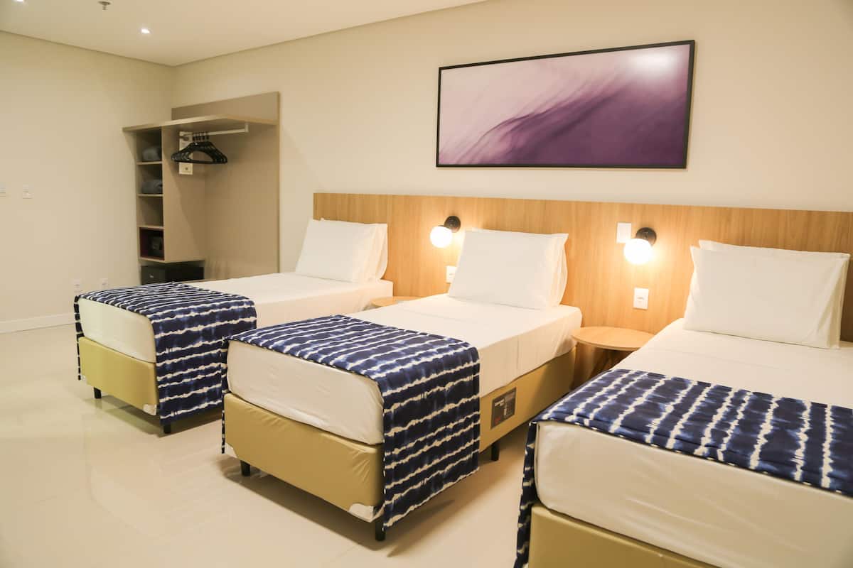 Quarto de hotel com três camas, cada uma com colcha de padrão azul e branco, cabeceira de madeira e luminária fixada na parede.
