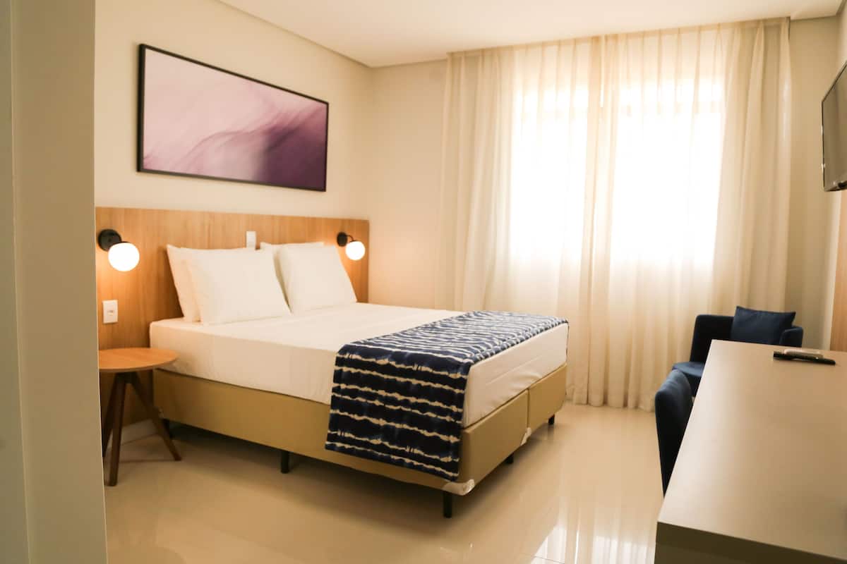 Quarto de hotel com cama, mesas de cabeceira, escrivaninha, cadeira, televisão e janela com cortinas.