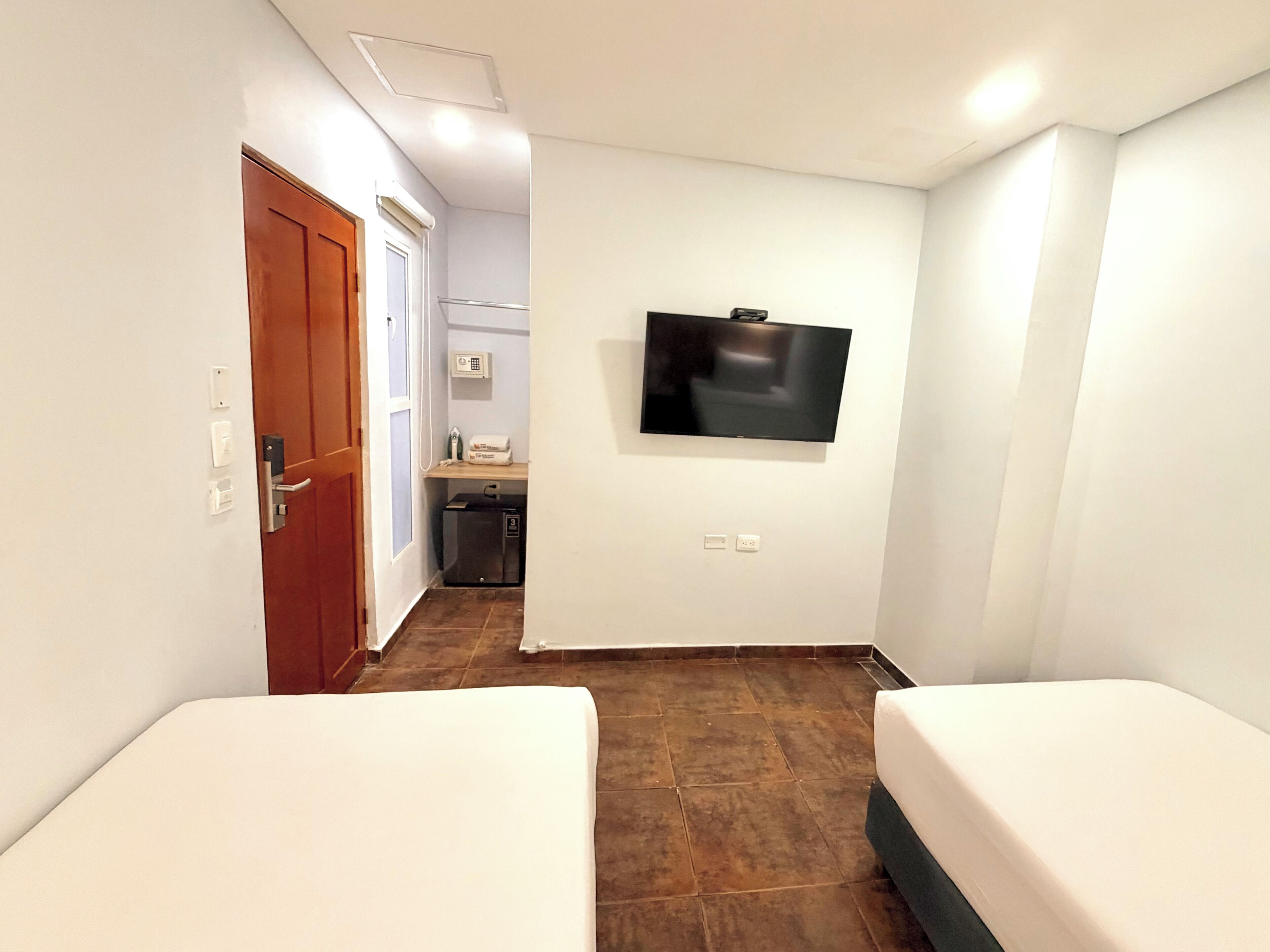 Quarto pequeno com duas camas, uma televisão na parede, uma porta e uma pequena cozinha.