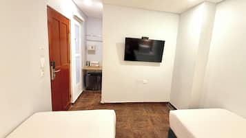Quarto pequeno com duas camas, uma televisão na parede, uma porta e uma pequena cozinha.