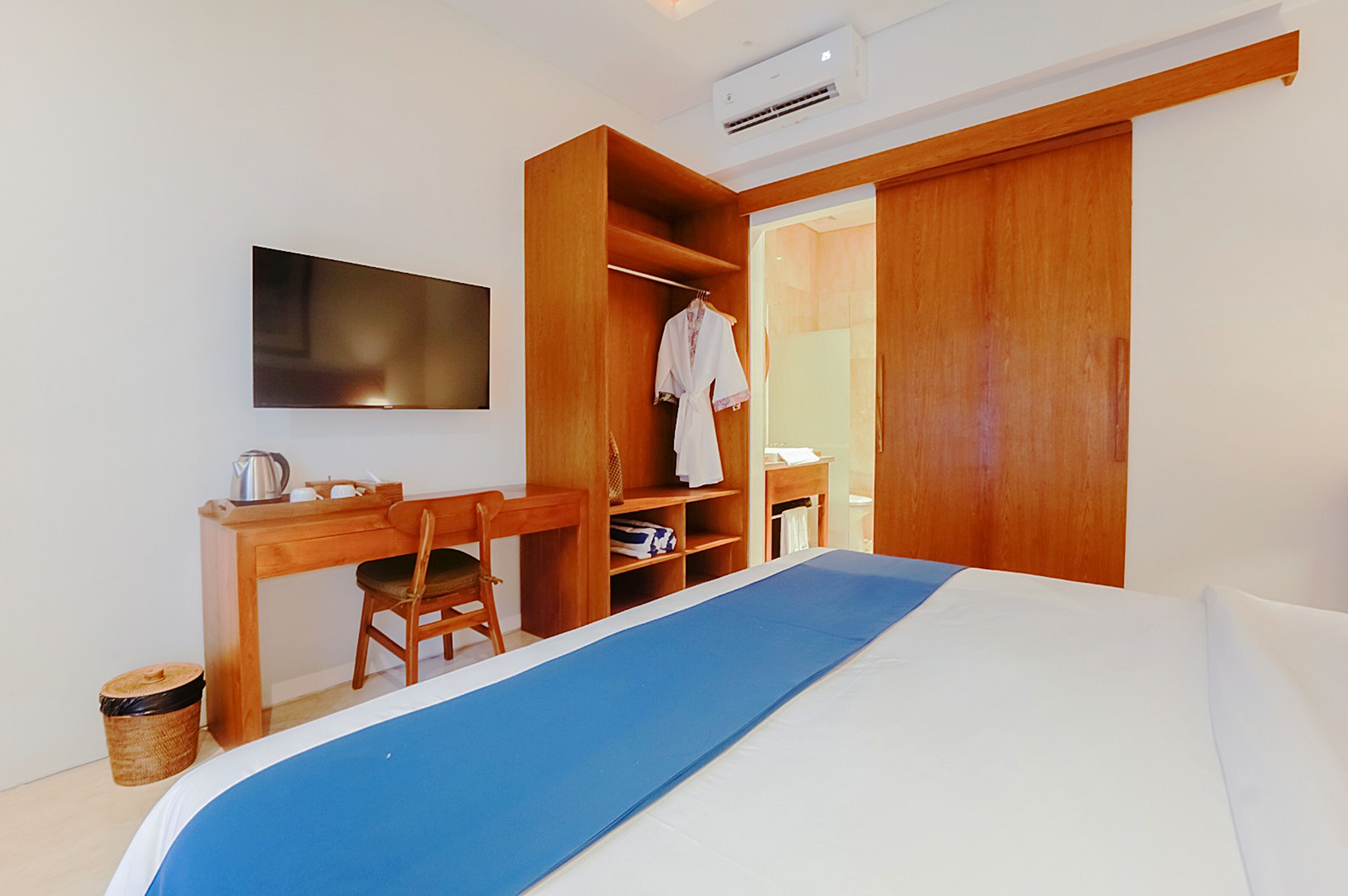 Quarto de hotel com cama, escrivaninha com cadeira, televisão, guarda-roupa e um pequeno cesto de lixo.