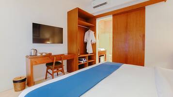 Quarto de hotel com cama, escrivaninha com cadeira, televisão, guarda-roupa e um pequeno cesto de lixo.
