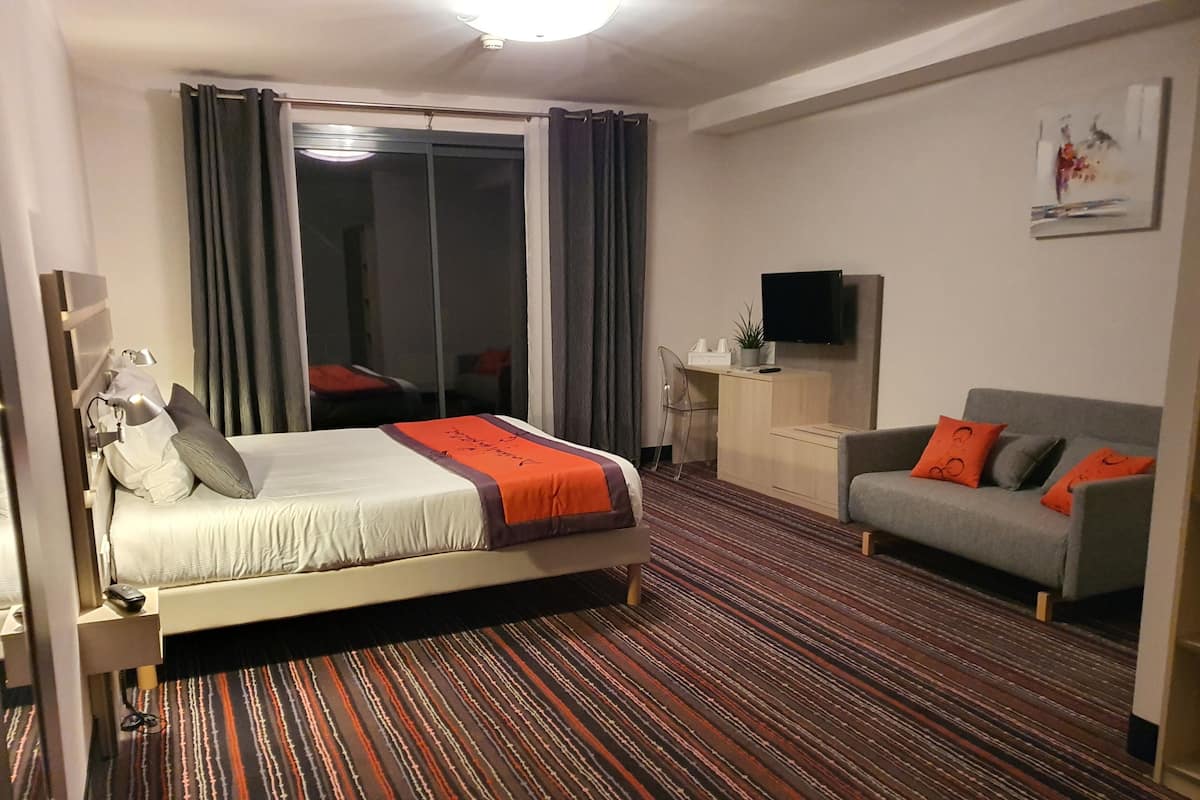 Quarto de hotel com cama, sofá, escrivaninha com televisão e janela com cortinas.