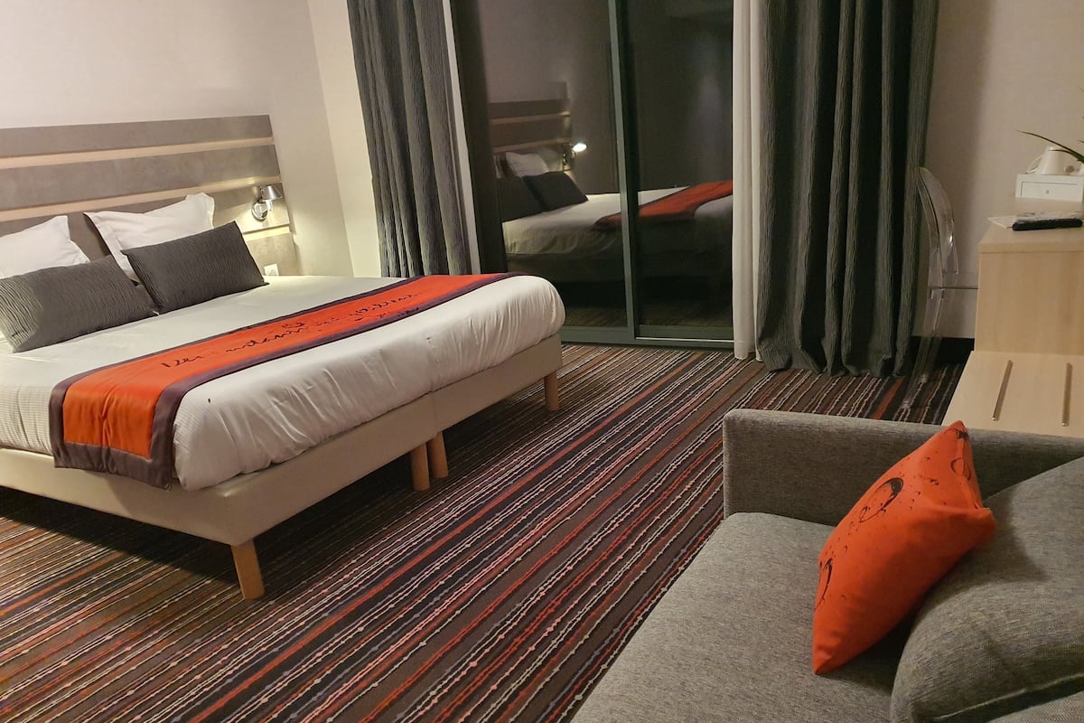 Quarto de hotel com cama, sofá, mesa de cabeceira e uma porta de correr para uma varanda.