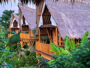 Um hotel no estilo de bangalô, com telhados de palha e varandas de madeira, rodeado por vegetação tropical exuberante.