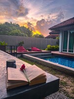 Ambiente externo tranquilo com piscina, um livro, uma rosa e uma caneca sobre uma mesa.